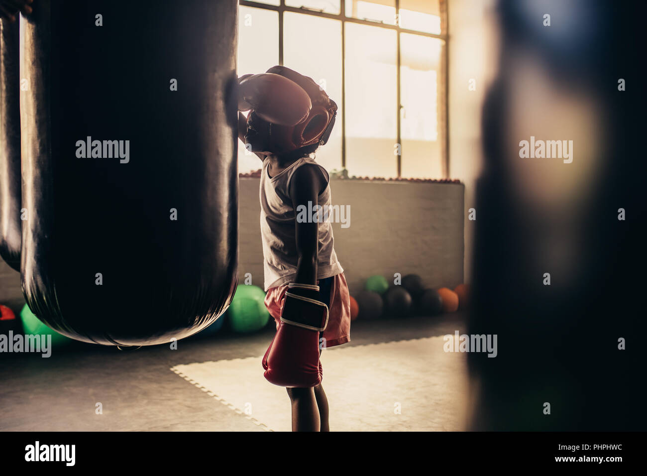 Tired kid in boxing gloves and headgear leaning against a punching bag ...