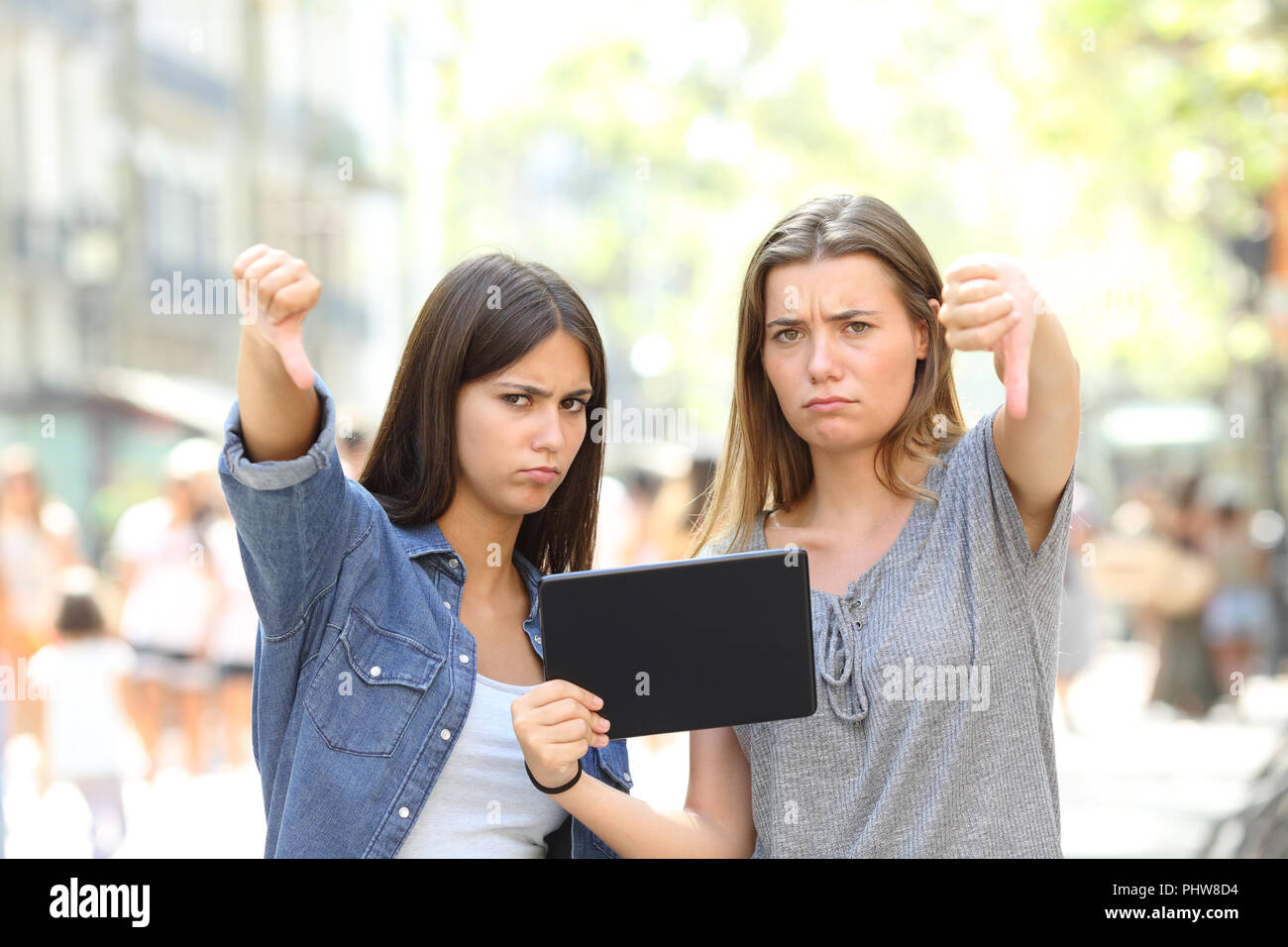 Two angry friends holding a tablet gesturing thumbs down in the street ...