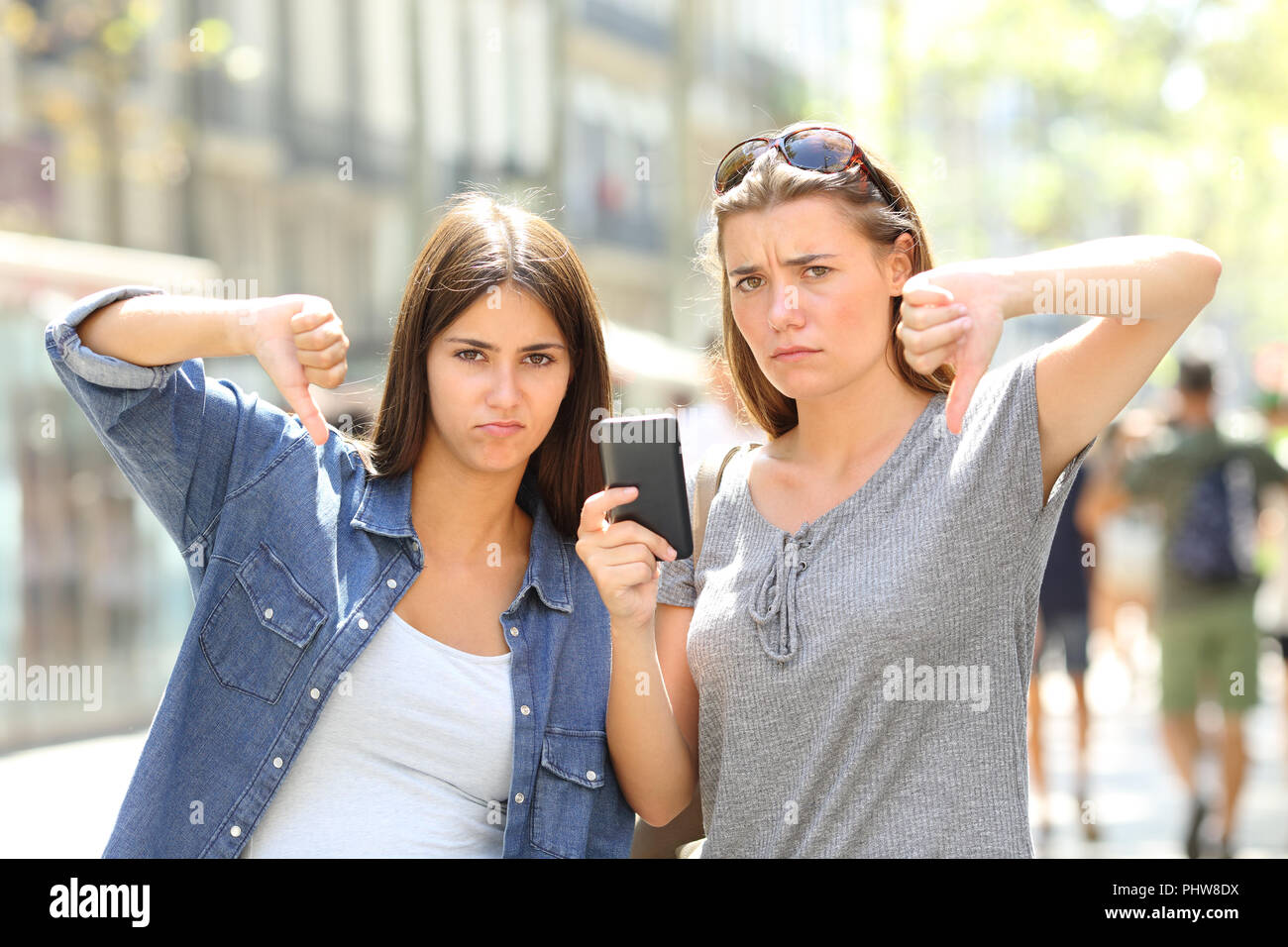 Two angry friends holding a smart phone with thumbs down in the street ...