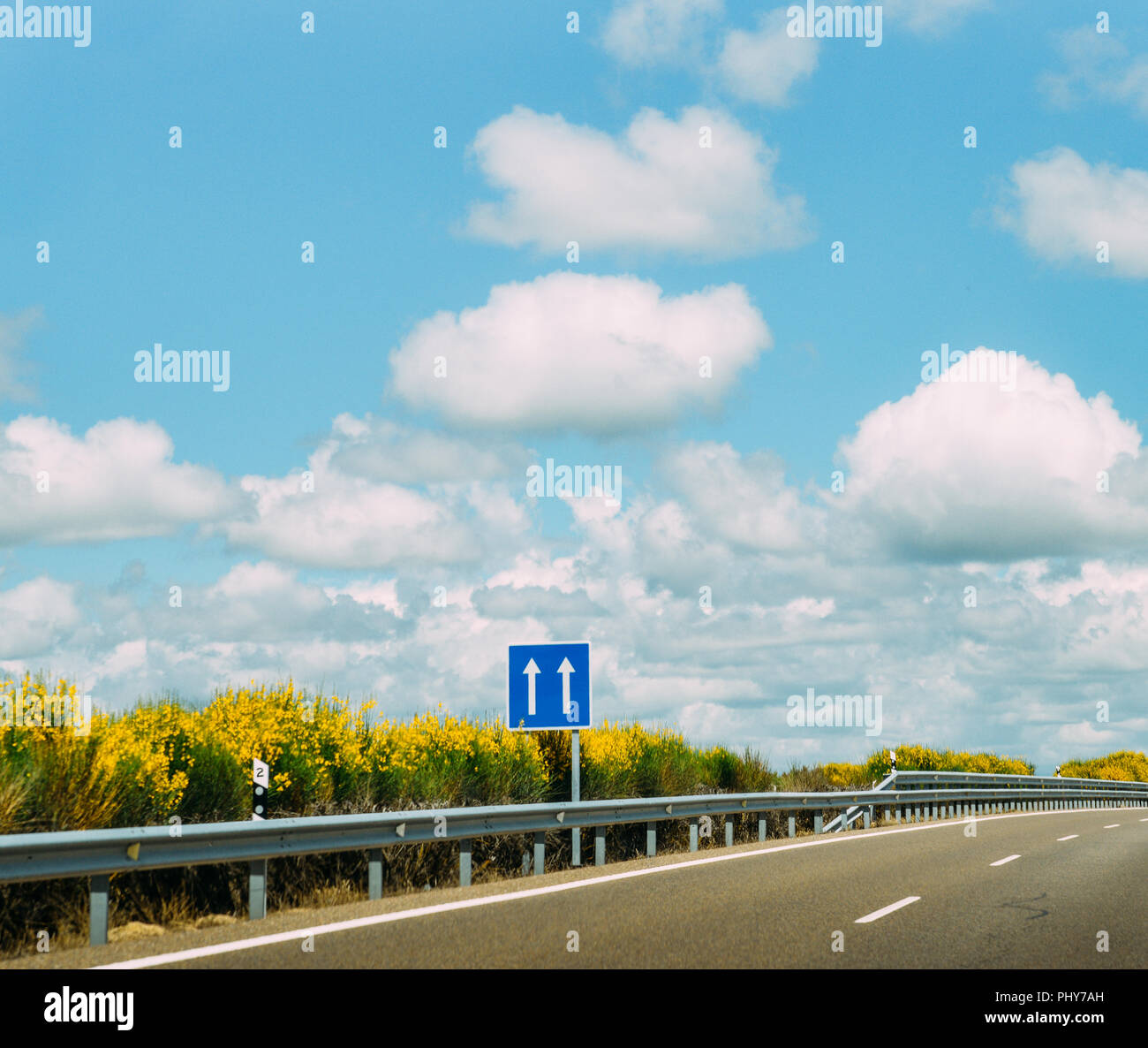 Blue sign pointing up at empty highway with blue sky and fluffly clouds ...