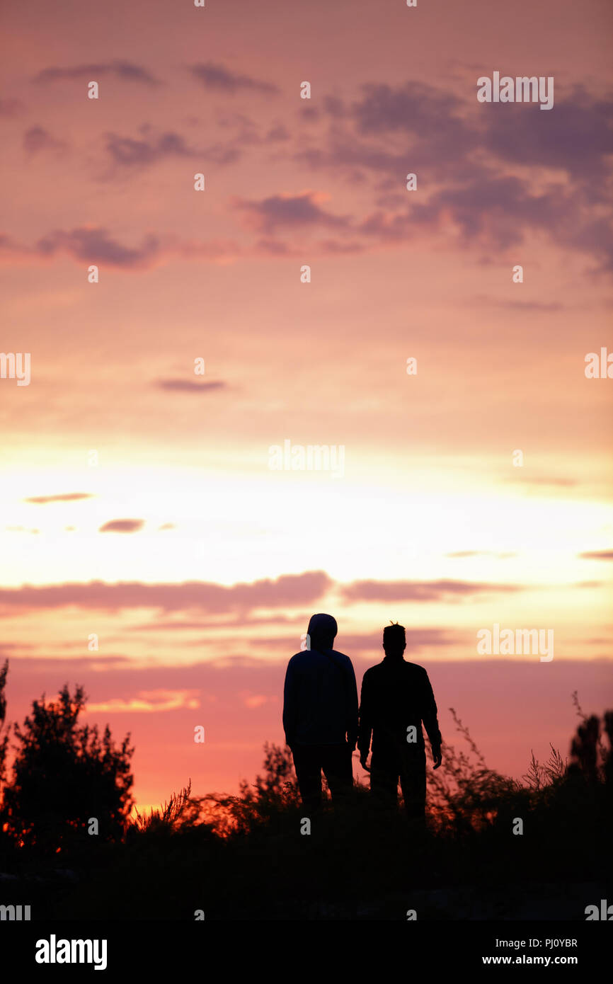Couple of young gay men standing together staring at the sun at sunet ...