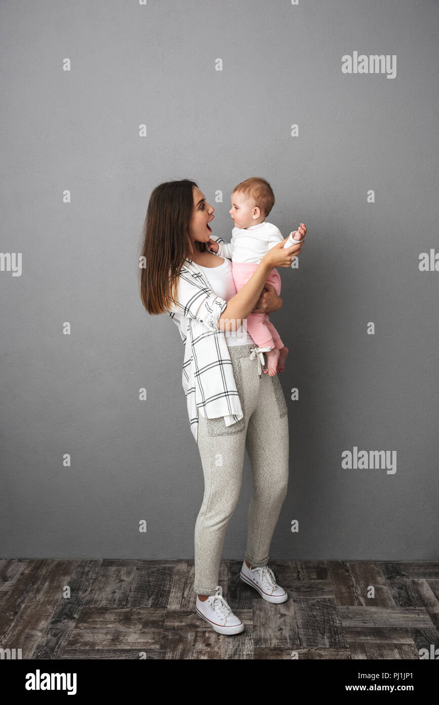 Full length portrait of a cheerful young mother holding her little baby girl over gray ...