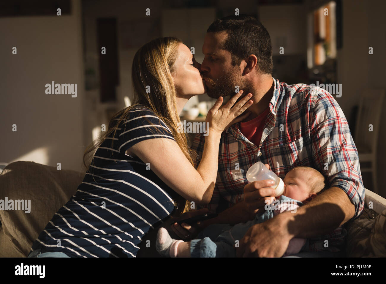 Parents kissing each other while father feeding to baby on sofa Stock ...