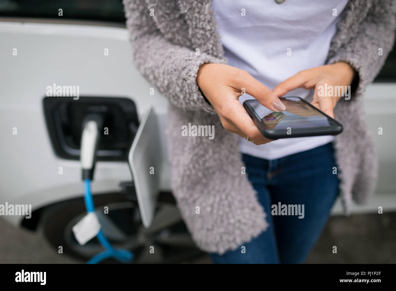 Woman using mobile phone while charging electric car at charging ...