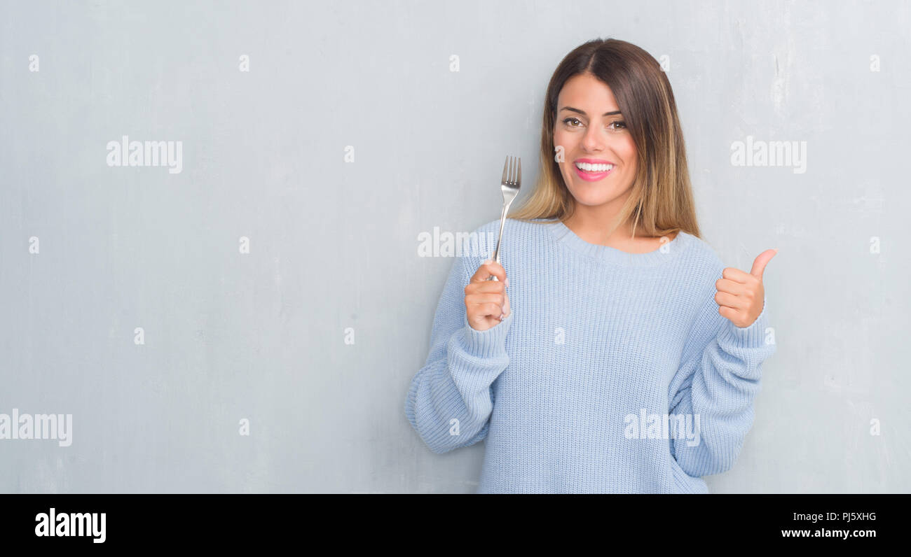 Young adult woman over grey grunge wall holding silver fork happy with ...