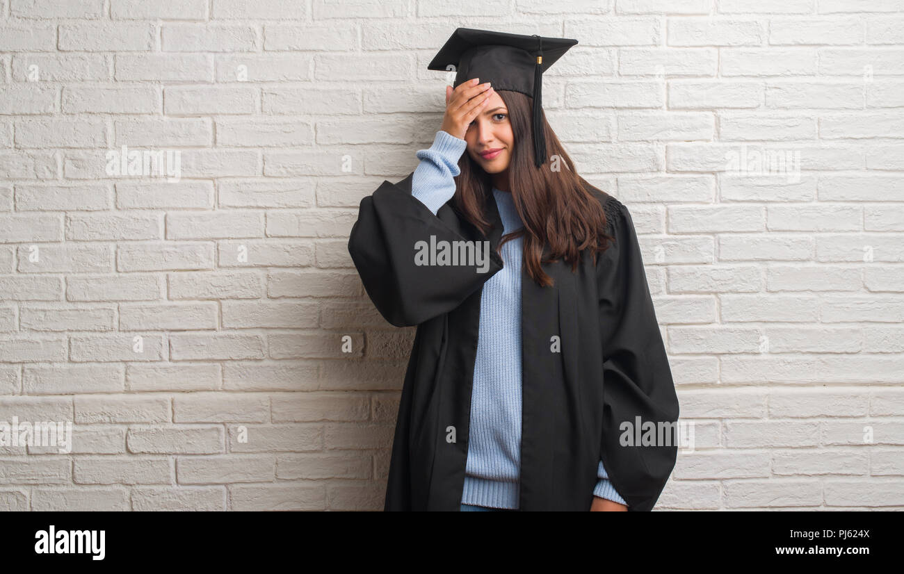 Young brunette woman standing over white brick wall wearing graduate ...