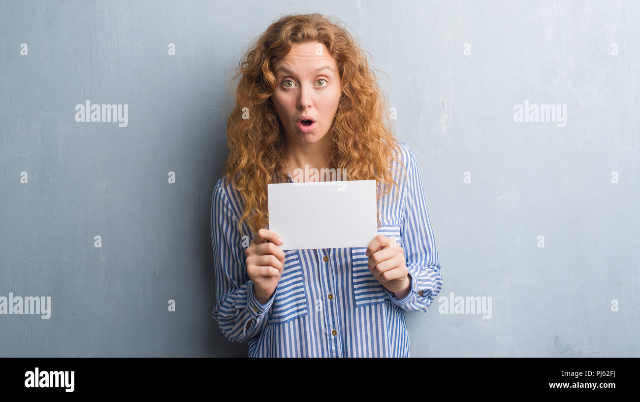 Young redhead woman over grey grunge wall holding blank card scared in ...