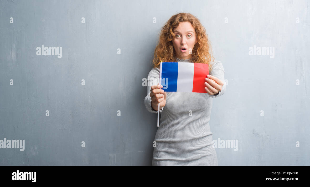 Young redhead woman over grey grunge wall holding flag of France scared ...