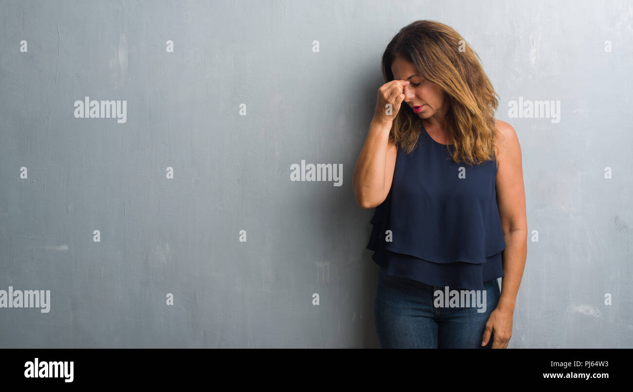 Middle age hispanic woman standing over grey grunge wall tired rubbing ...