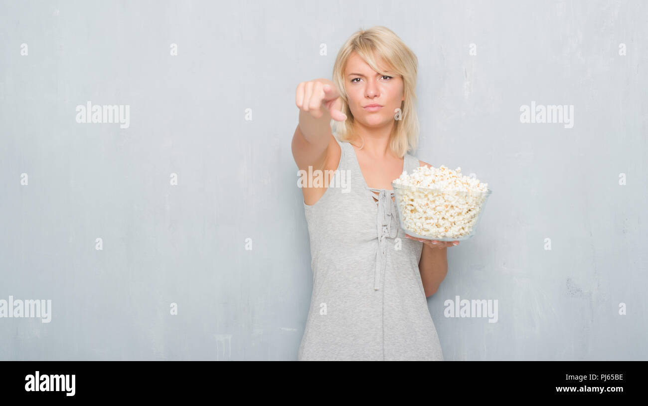 Adult caucasian woman over grunge grey wall eating pop corn pointing ...