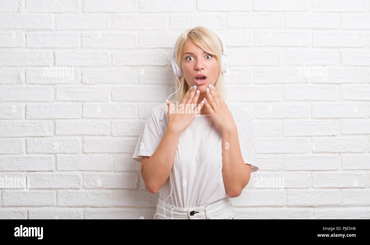Adult caucasian woman over white brick wall wearing headphones afraid ...