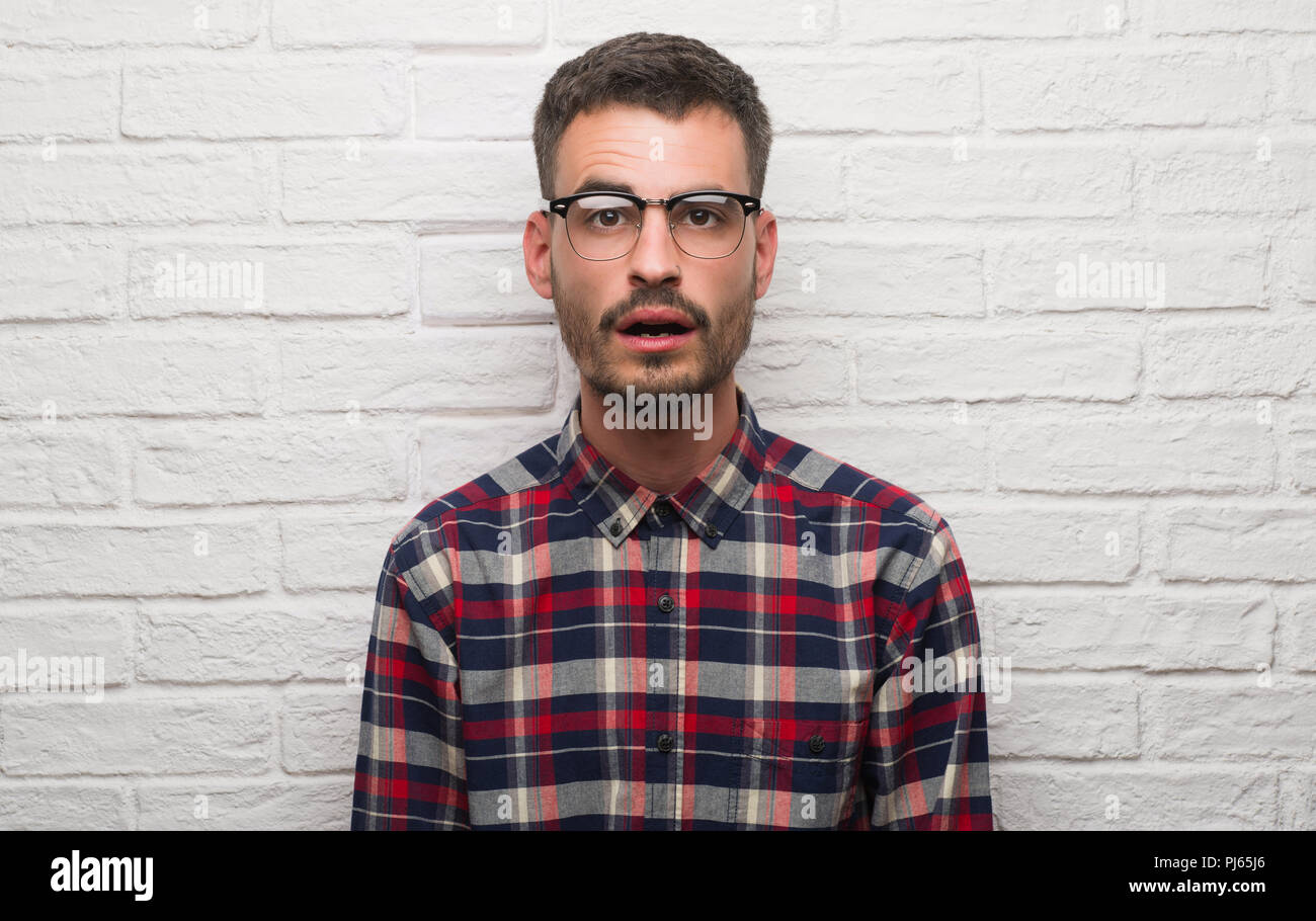 Young adult man standing over white brick wall scared in shock with a ...