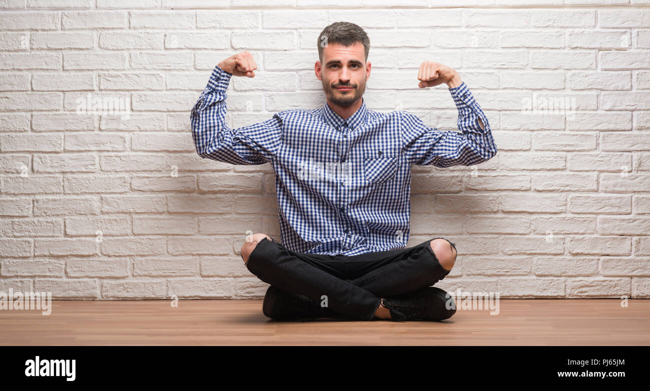 Young adult man sitting over white brick wall showing arms muscles ...