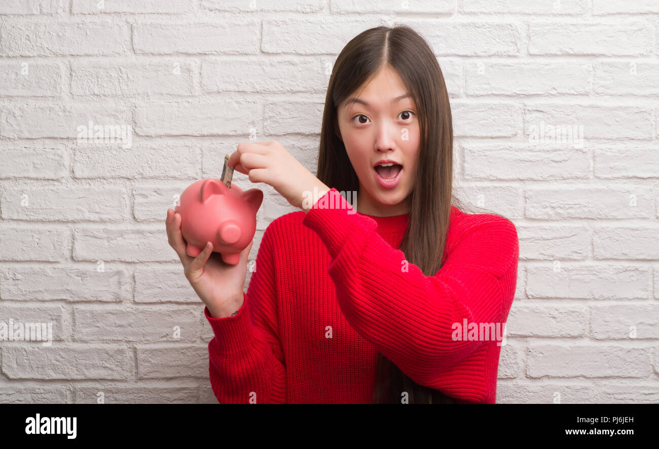 Young Chinese woman over brick wall holding piggy bank scared in shock ...