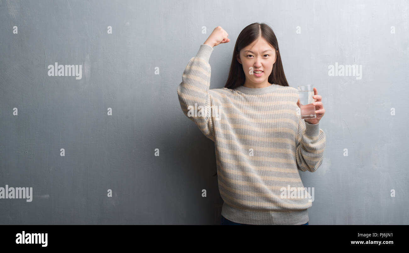 Young Chinese woman over grey wall drinking water annoyed and ...