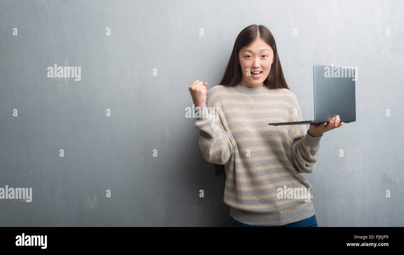 Young Chinese woman over grey wall using computer laptop screaming ... Young Chinese woman over grey wall using computer laptop screaming ...