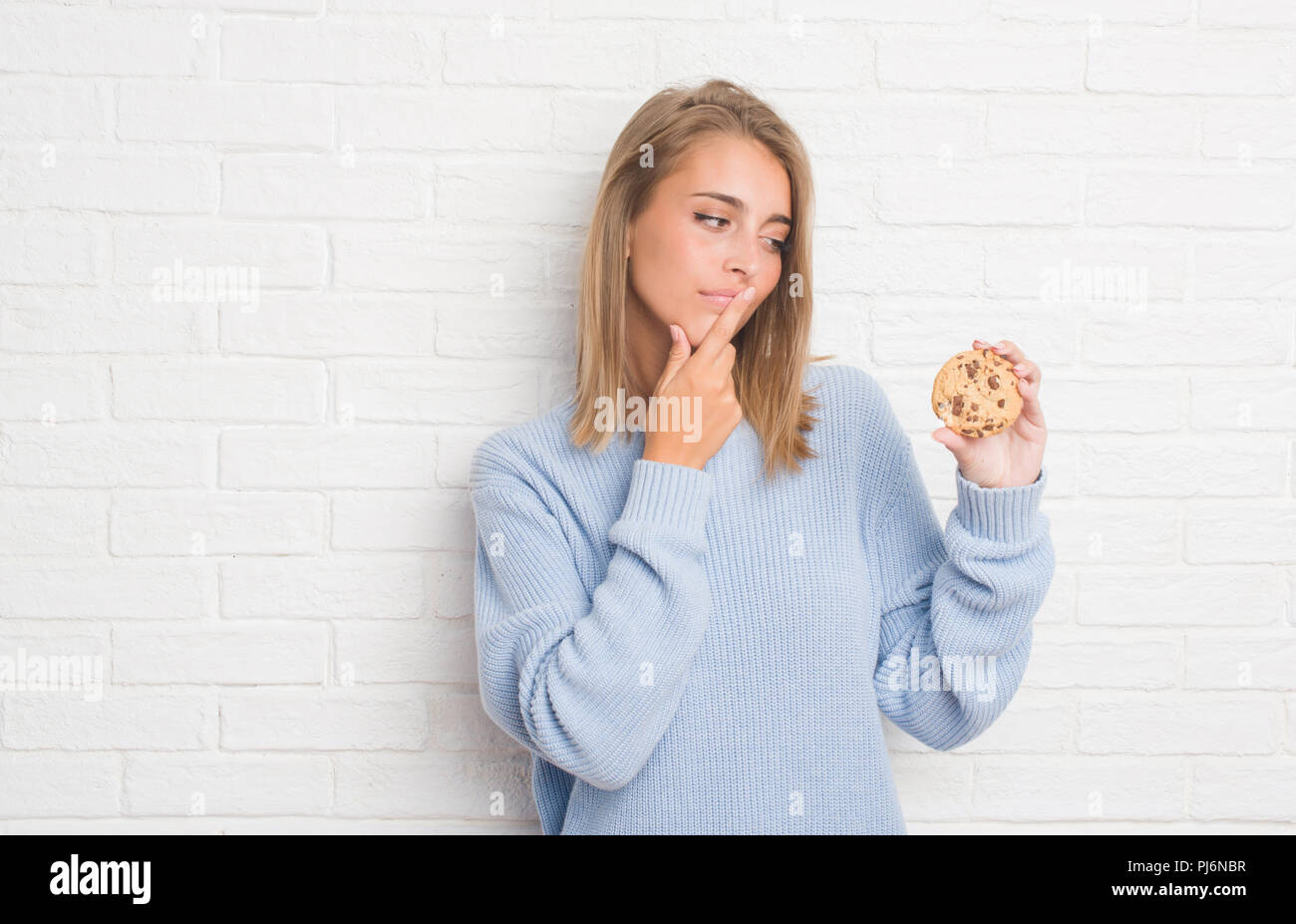 Beautiful young woman over white brick wall eating chocolate chip cooky ...