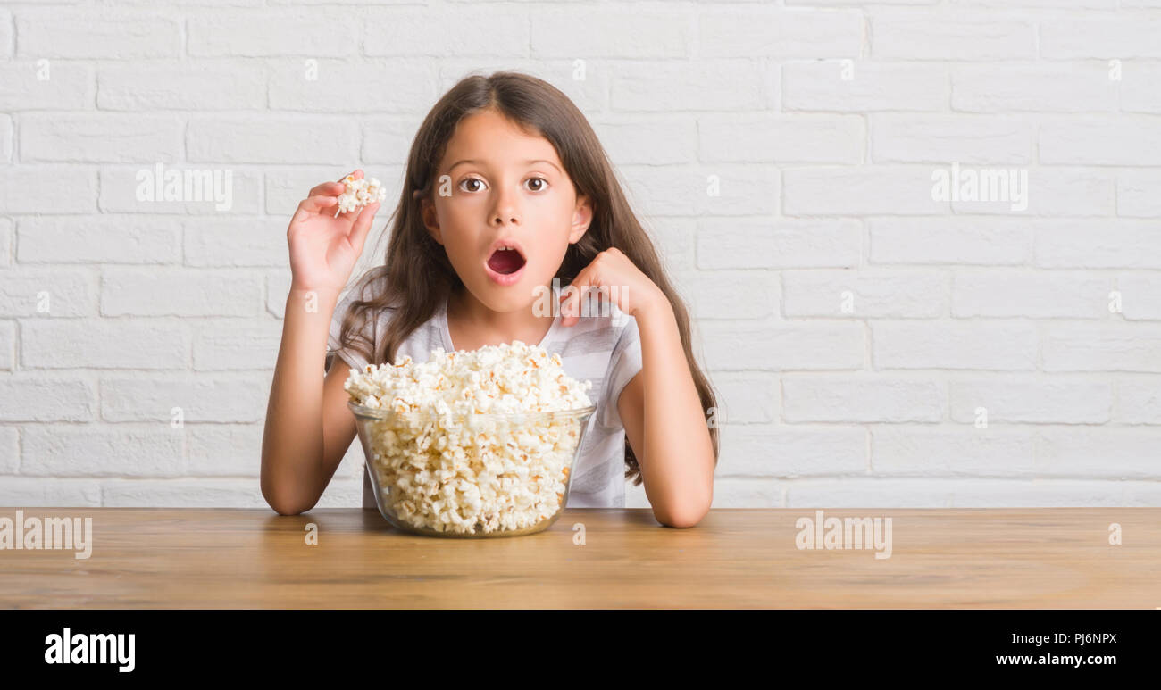 Young hispanic kid sitting on the table eating pop corn scared in shock ...