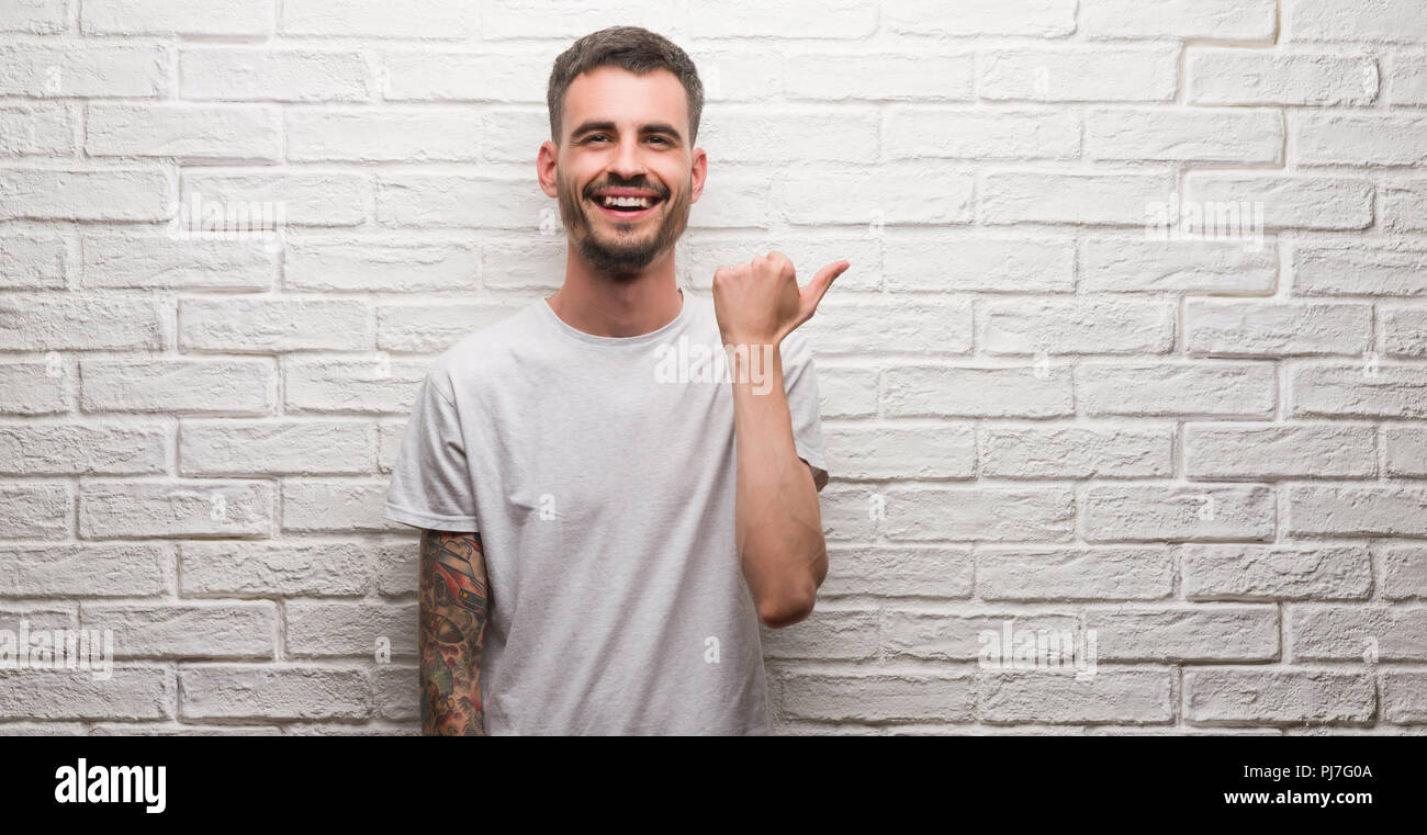 Young adult man standing over white brick wall pointing and showing ...