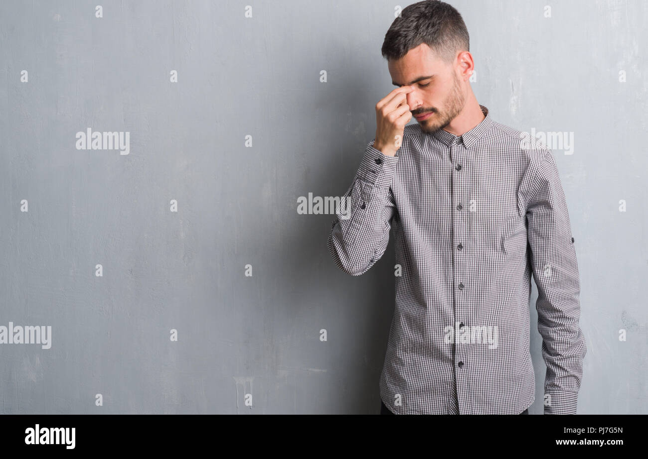 Young adult man standing over grey grunge wall tired rubbing nose and ...