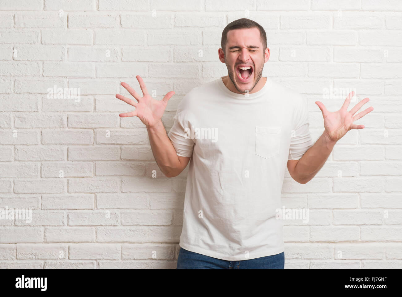 Young caucasian man standing over white brick wall celebrating mad and ...