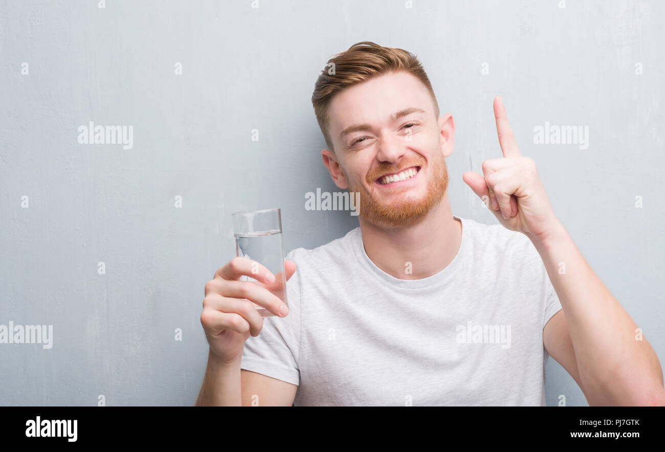 Young redhead man over grey grunge wall drinking water surprised with ...