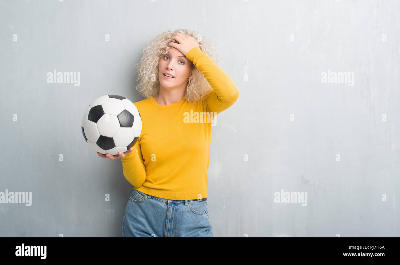 Young blonde woman over grunge grey background holding soccer football ...