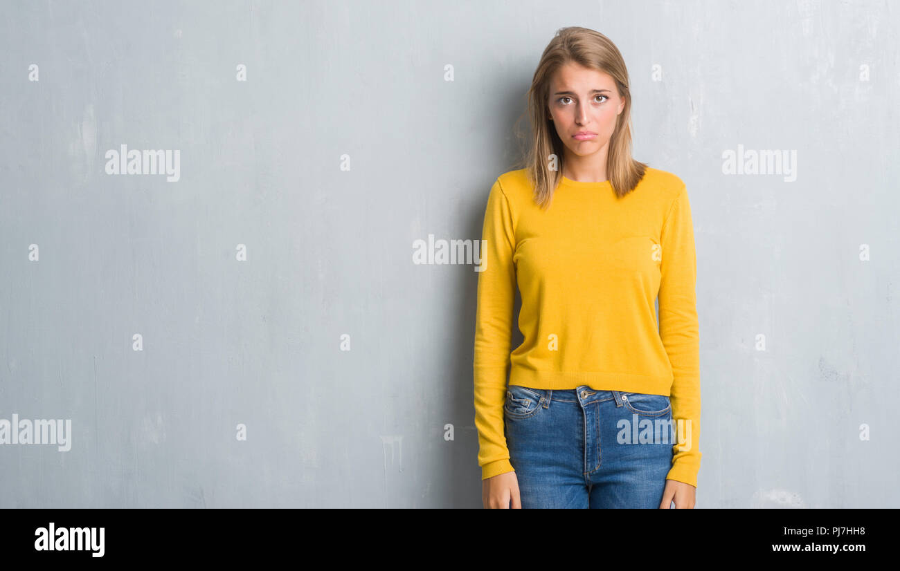Beautiful young woman standing over grunge grey wall depressed and ...