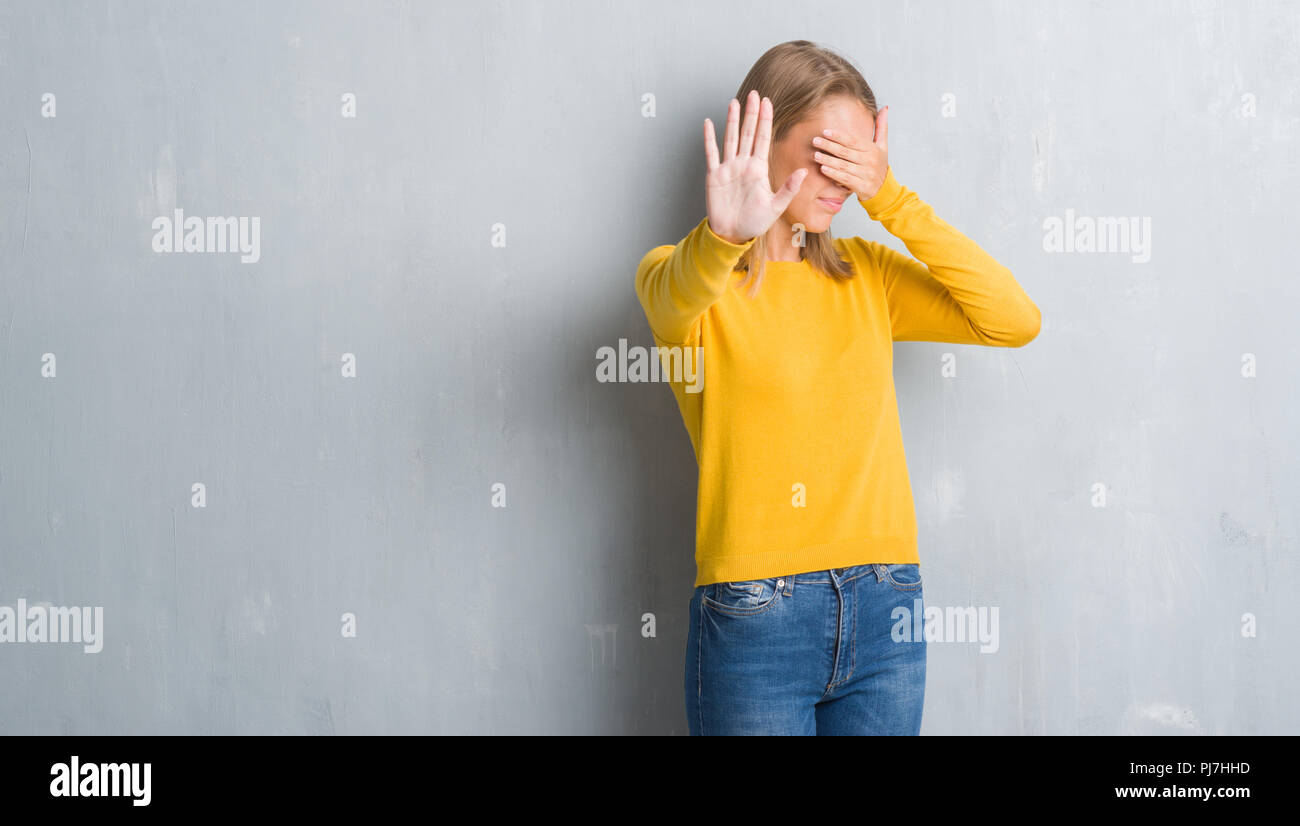Beautiful young woman standing over grunge grey wall covering eyes with ...