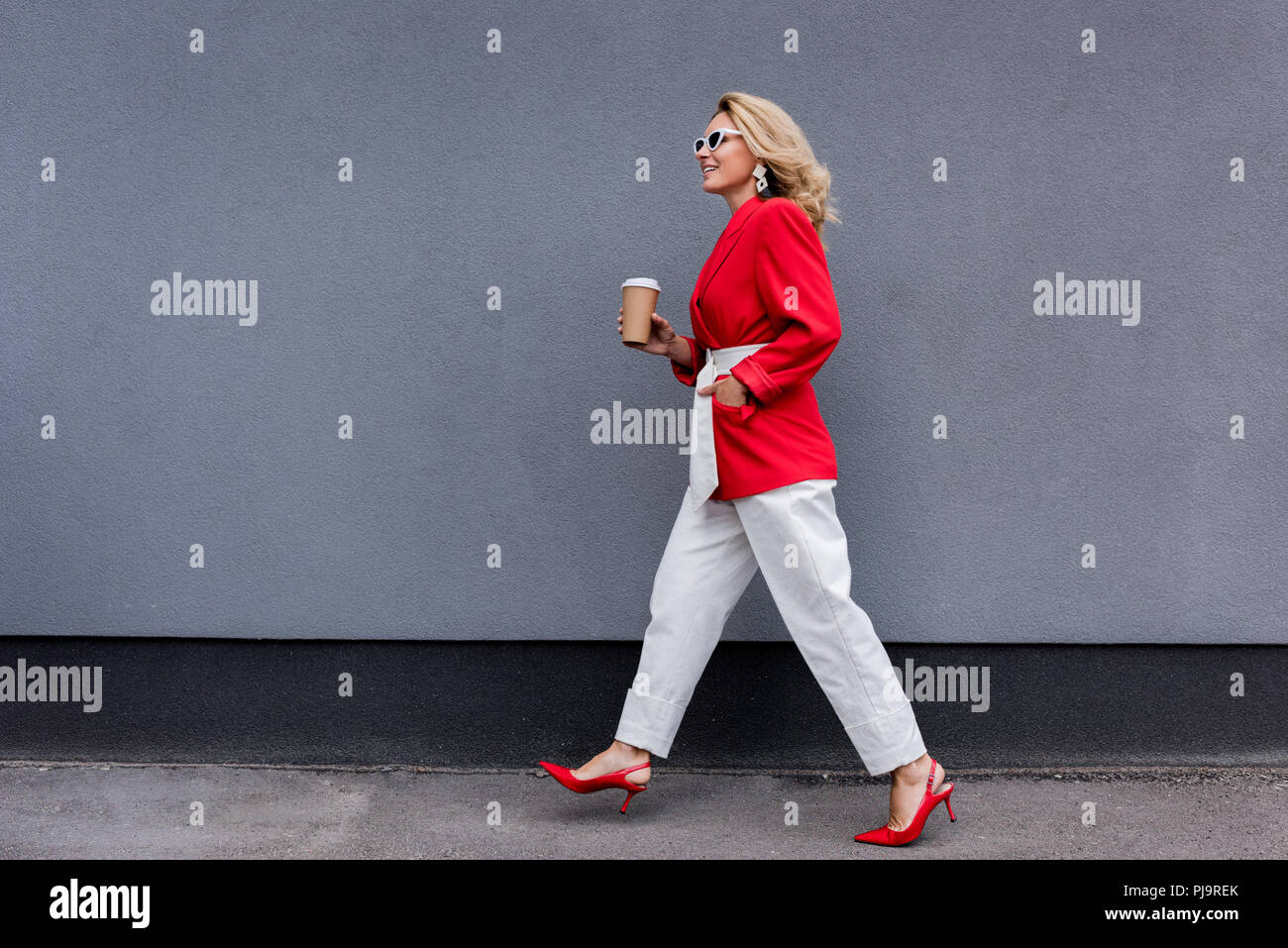 side view of attractive woman in red jacket walking with coffee in ...