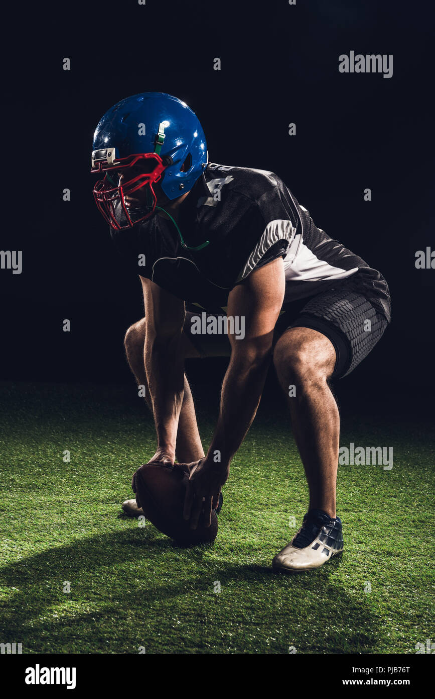american football player standing squats on grass with ball between ...