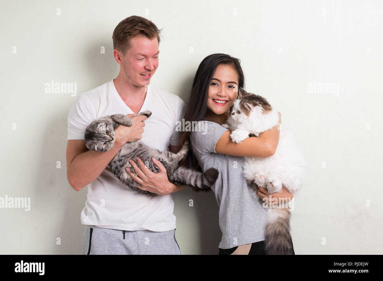 Young happy multi-ethnic couple holding two cats together against white ...