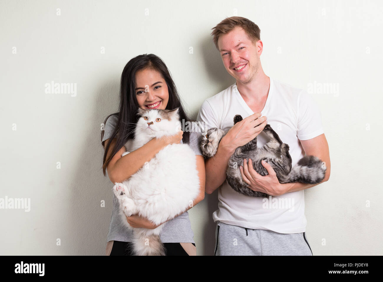 Young happy multi-ethnic couple holding two cats together against white ...