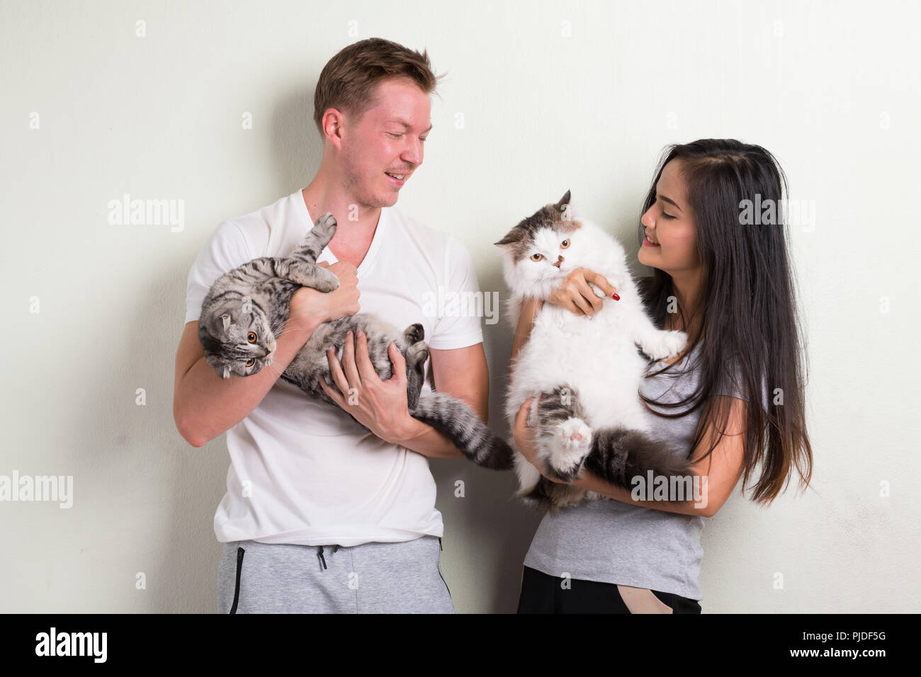 Young happy multi-ethnic couple holding two cats together against white ...