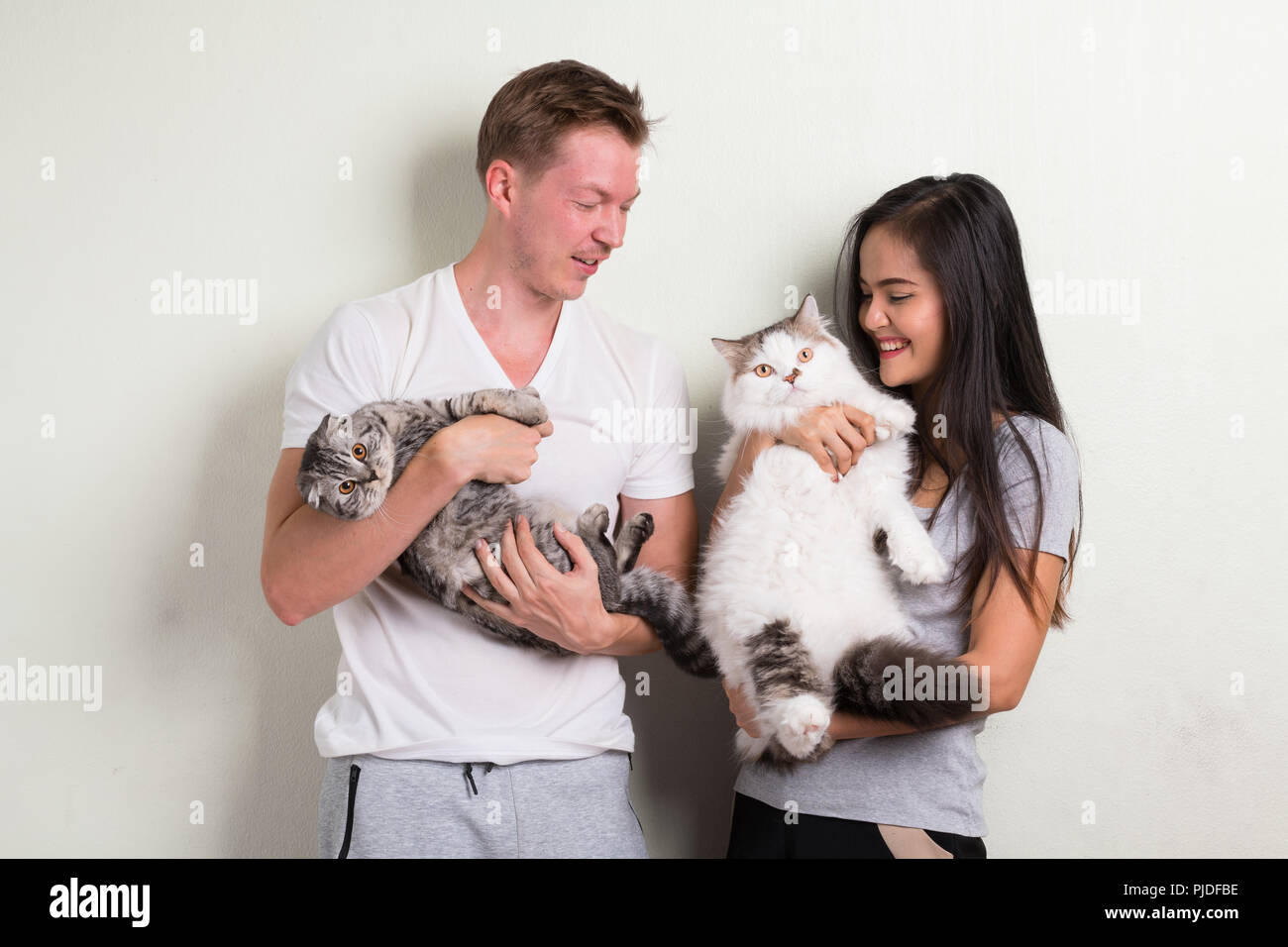 Young happy multi-ethnic couple holding two cats together against white ...