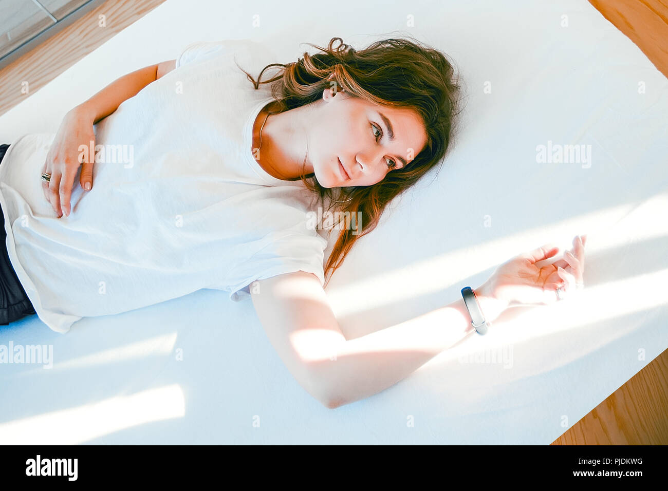 Young woman relaxing on bed, arm resting in ray of light, thoughtful ...