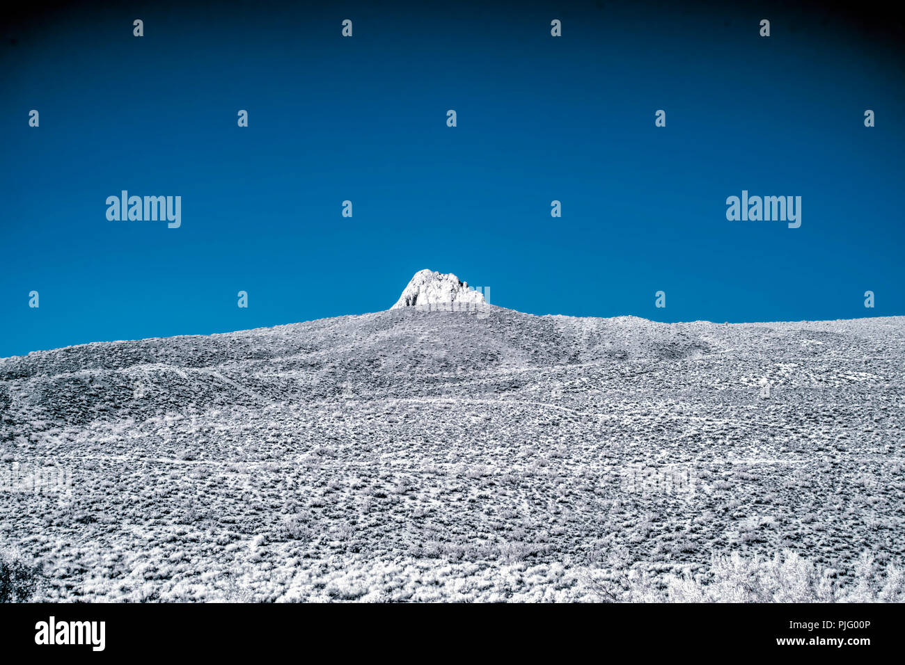 Rock formation peaking over desert hills under a blue sky. Black and ...
