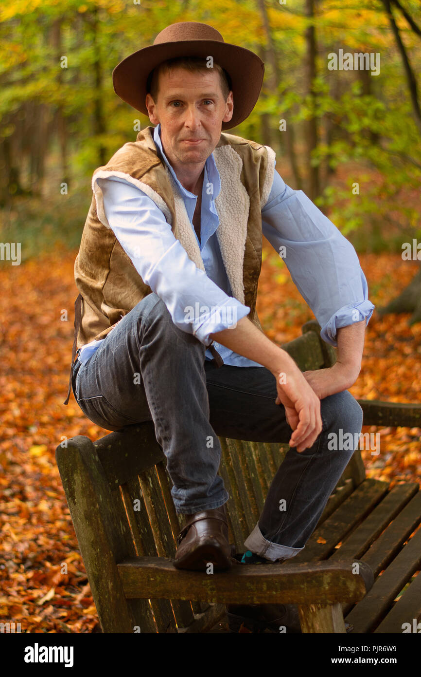 A man, dressed like a cowboy, takes a break sitting on a wooden bench ...