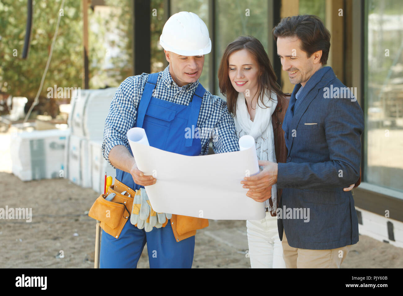 Worker shows house design plans to a young couple at construction site ...