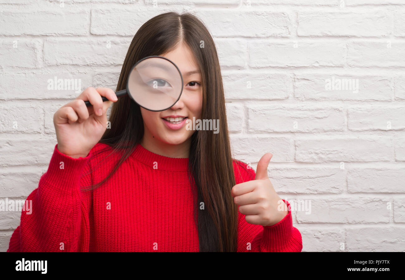 Young Chinese woman over brick wall looking through magnifying glass ...