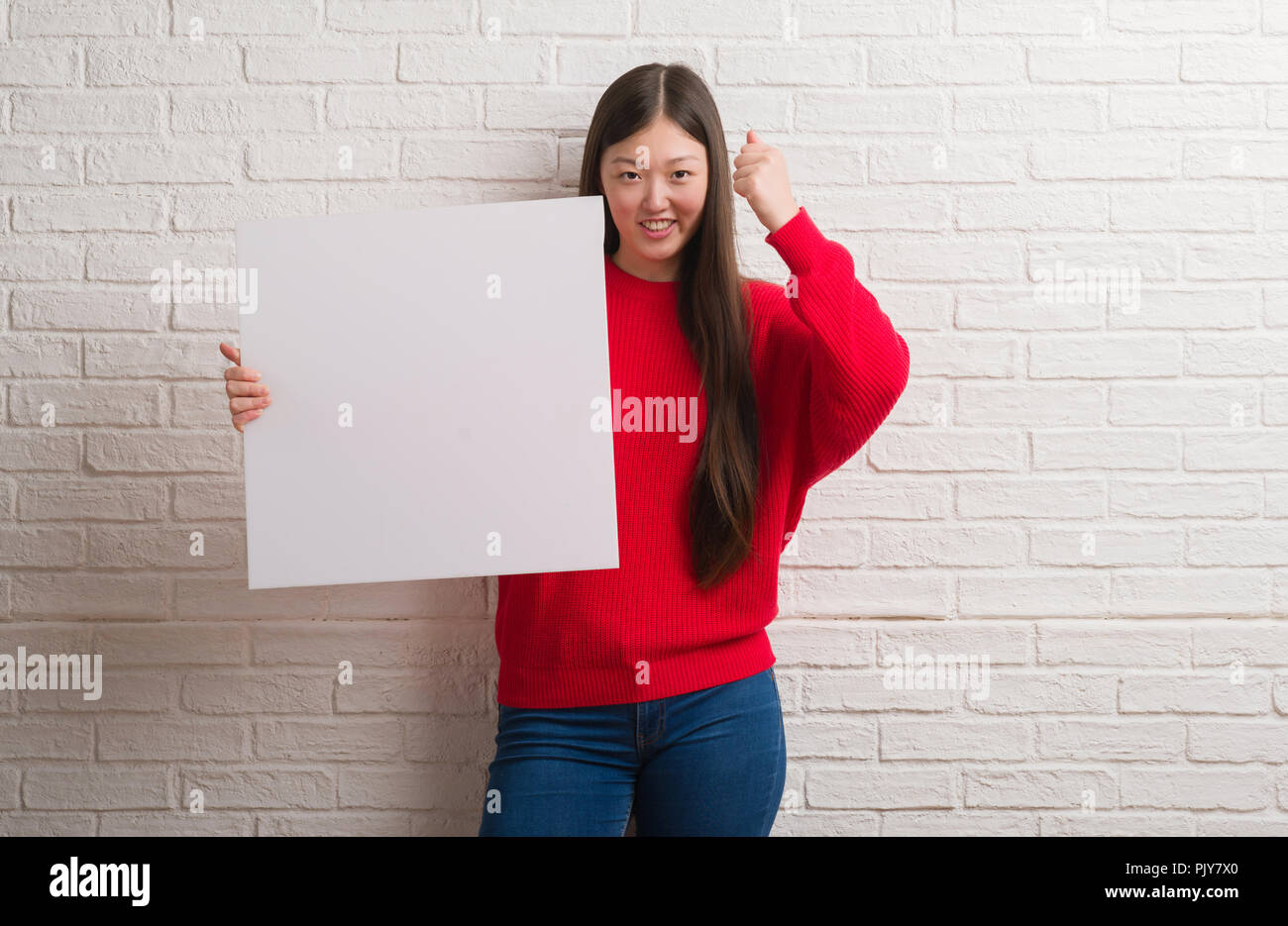 Young Chinese woman over brick wall holding banner annoyed and ...