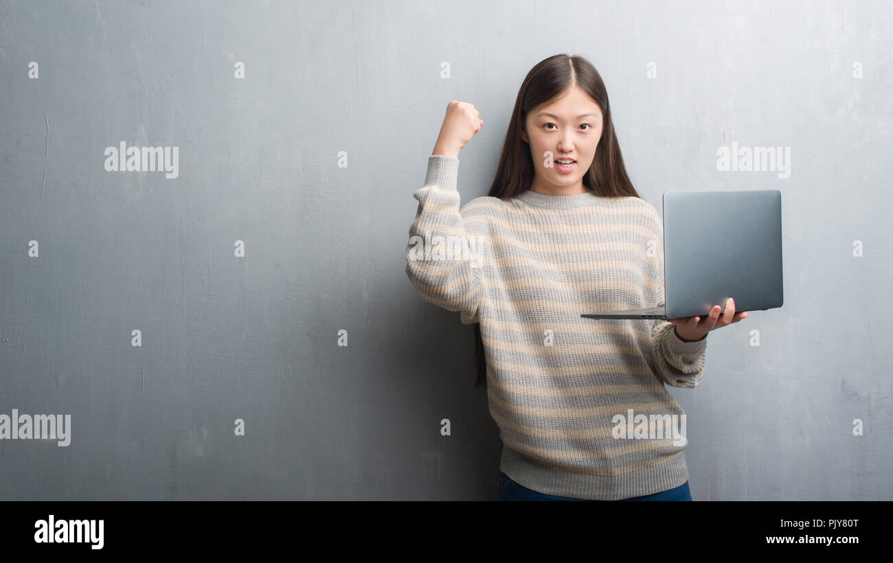 Young Chinese woman over grey wall using computer laptop annoyed and ...