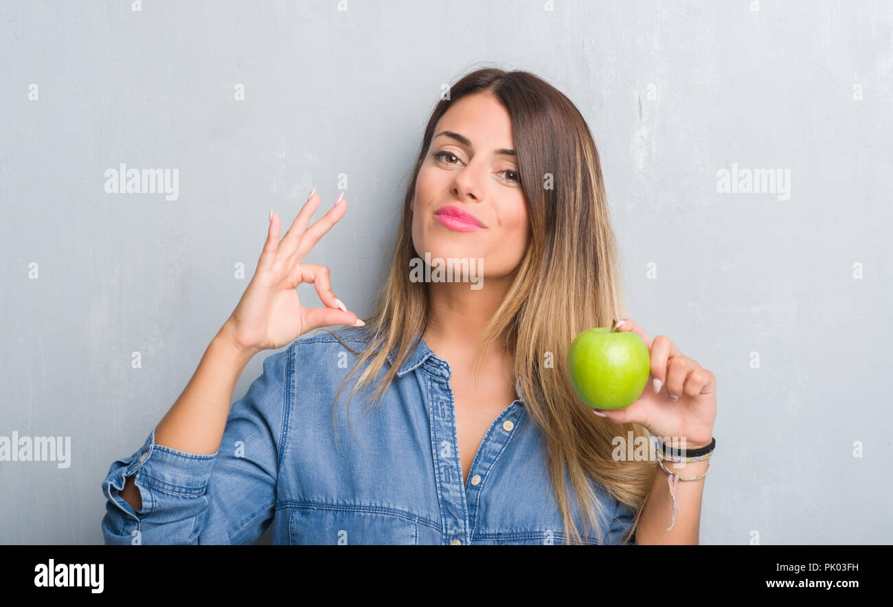 Young adult woman over grey grunge wall eating fresh green apple doing ...