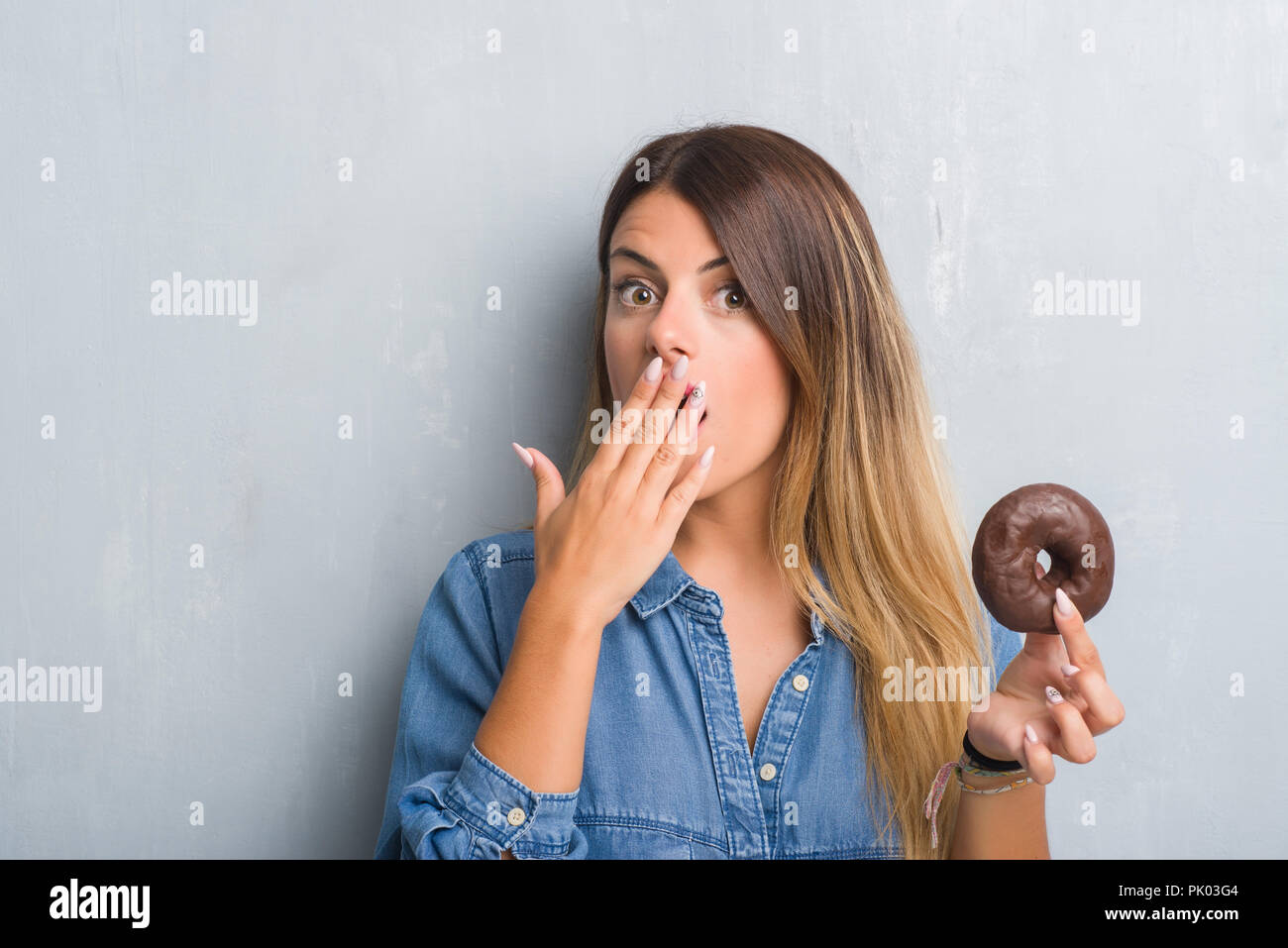 Young adult woman over grey grunge wall eating chocolate donut cover ...