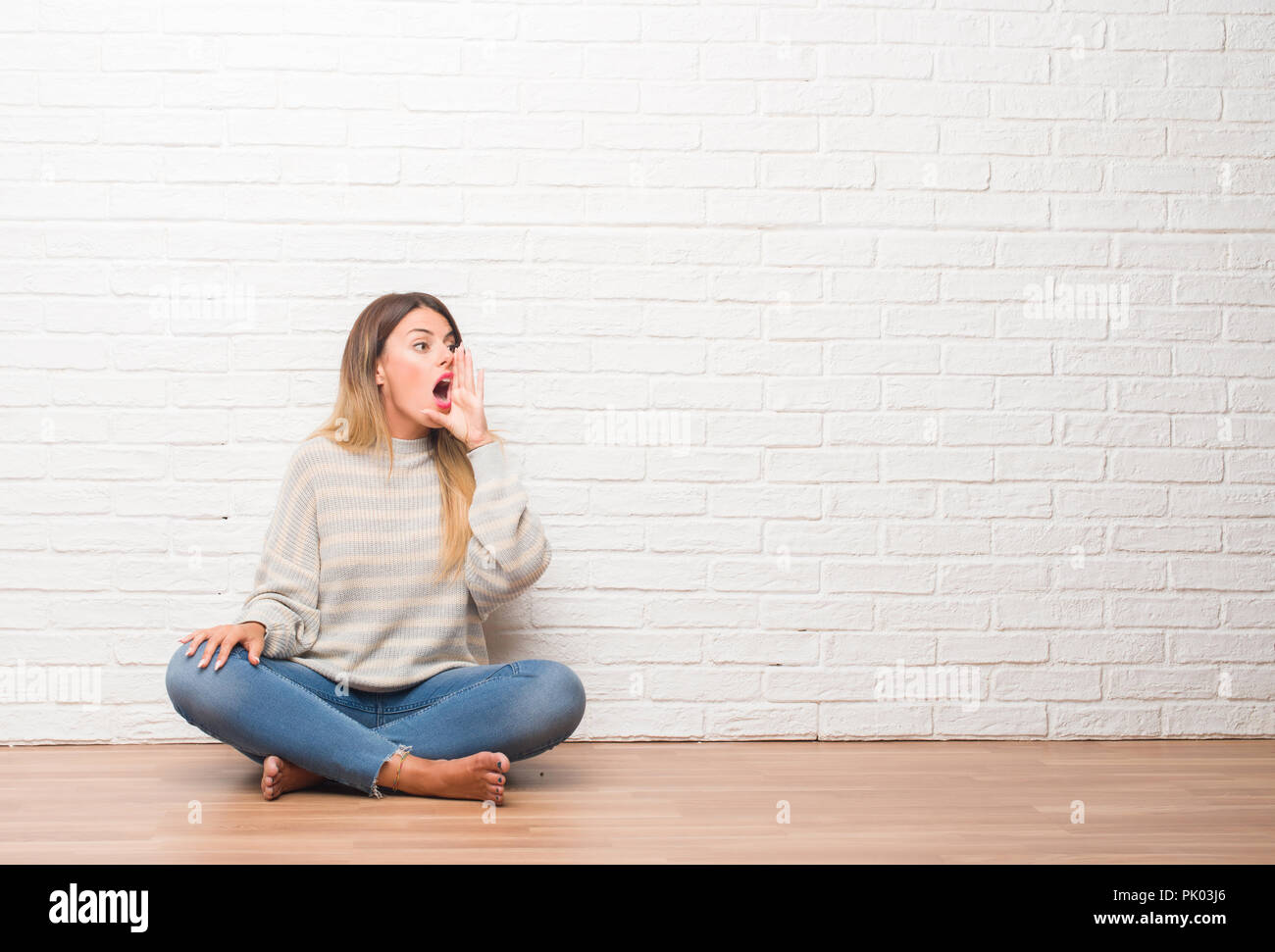Young adult woman sitting on the floor over white brick wall at home ...