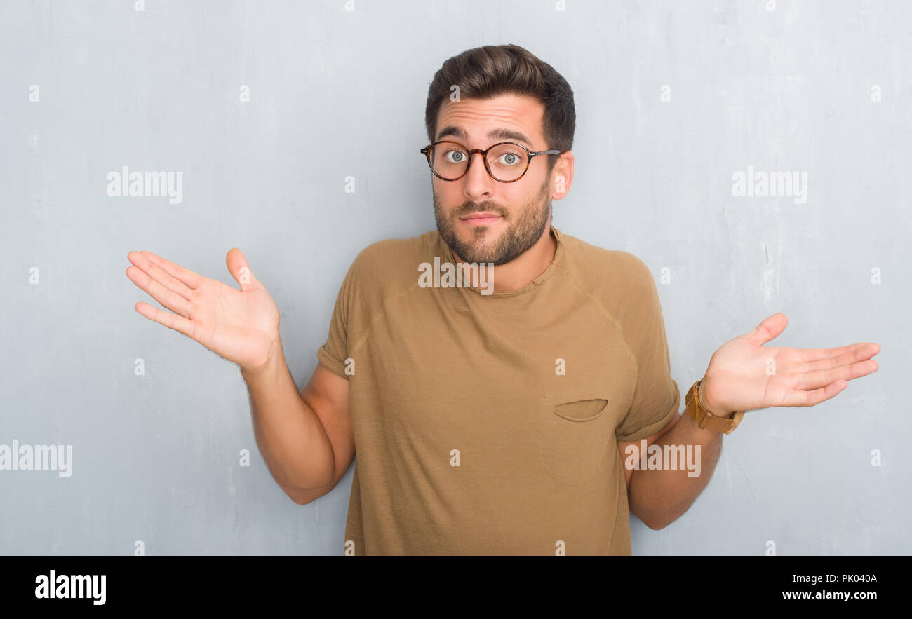 Handsome young man over grey grunge wall wearing glasses clueless and ...