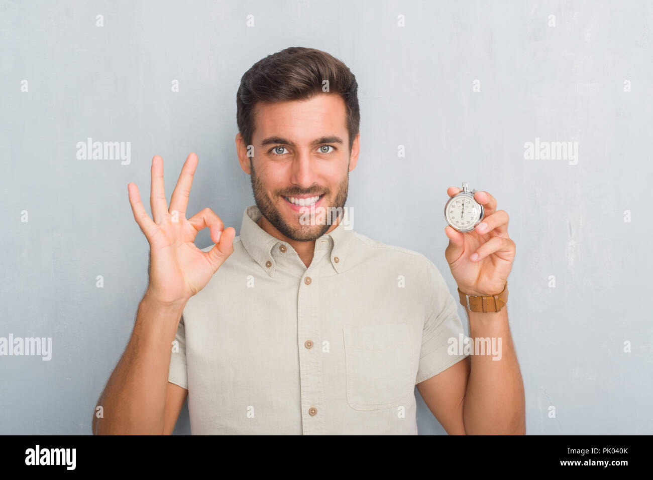 Handsome young man over grey grunge wall holding stopwatch doing ok ...