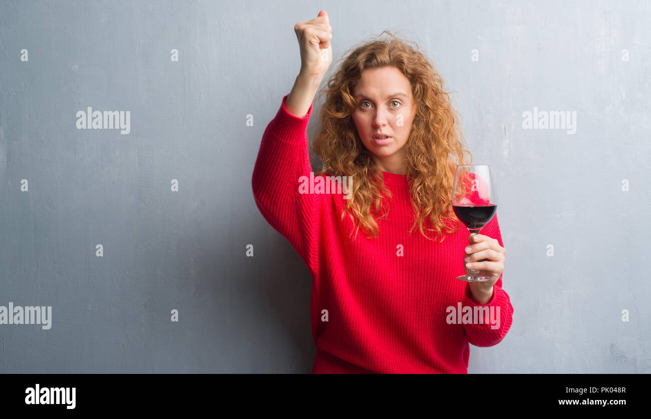 Young redhead woman over grey grunge wall drinking a glass of wine ...