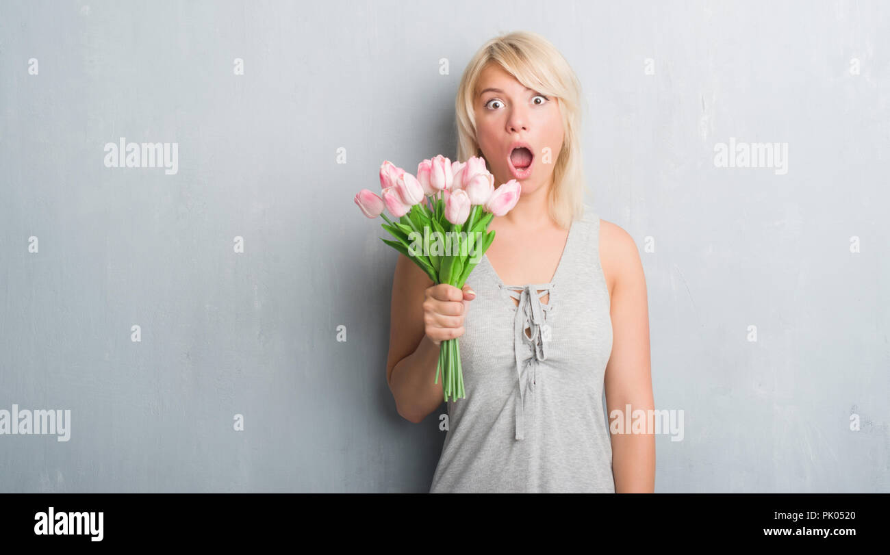 Caucasian adult woman over grey grunge wall holding pink flowers scared ...