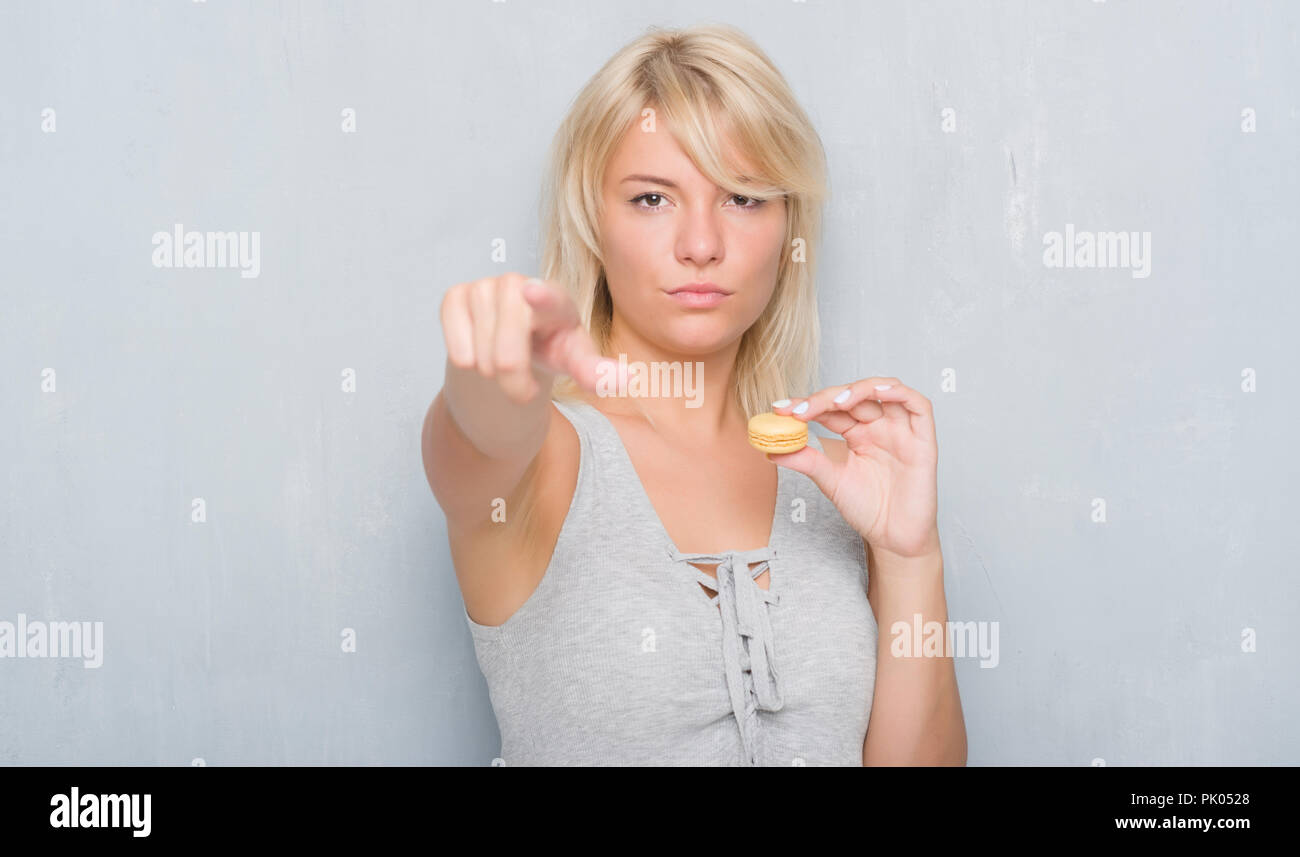 Adult caucasian woman over grunge grey wall eating macaron pointing ...